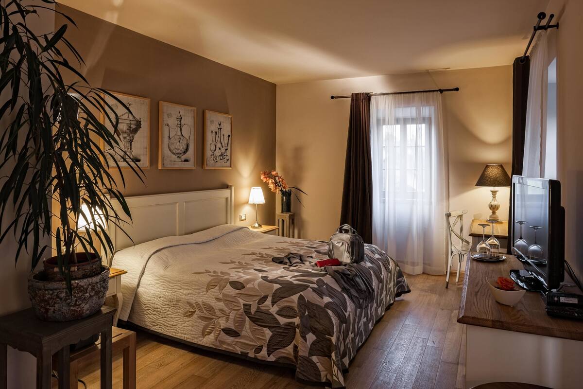 Quarto de hotel com cama, mesa de cabeceira, abajur, planta em vaso e televisão.