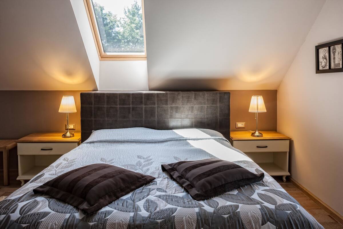 Quarto com teto inclinado, duas mesas de cabeceira com luminárias, cama com roupa de cama estampada e um claraboia.