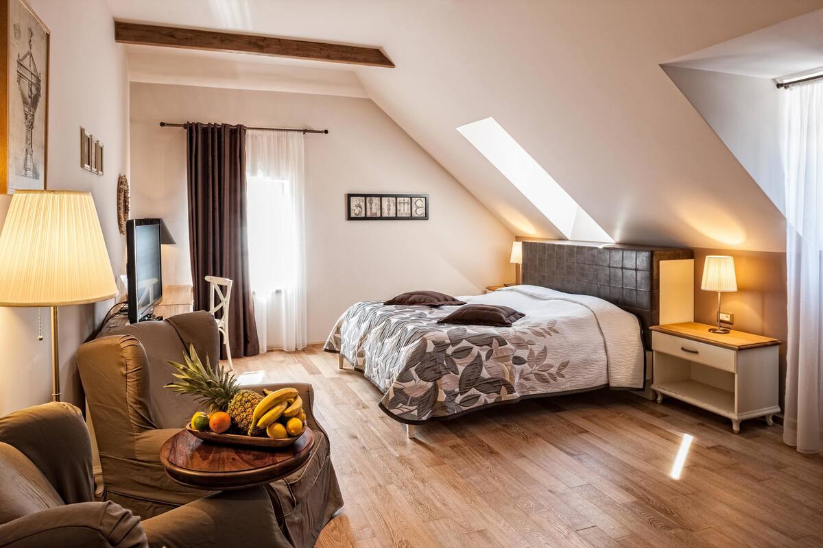 Um quarto com teto inclinado, uma cama com colcha estampada, uma mesa de cabeceira com abajur e uma fruteira na mesinha lateral.