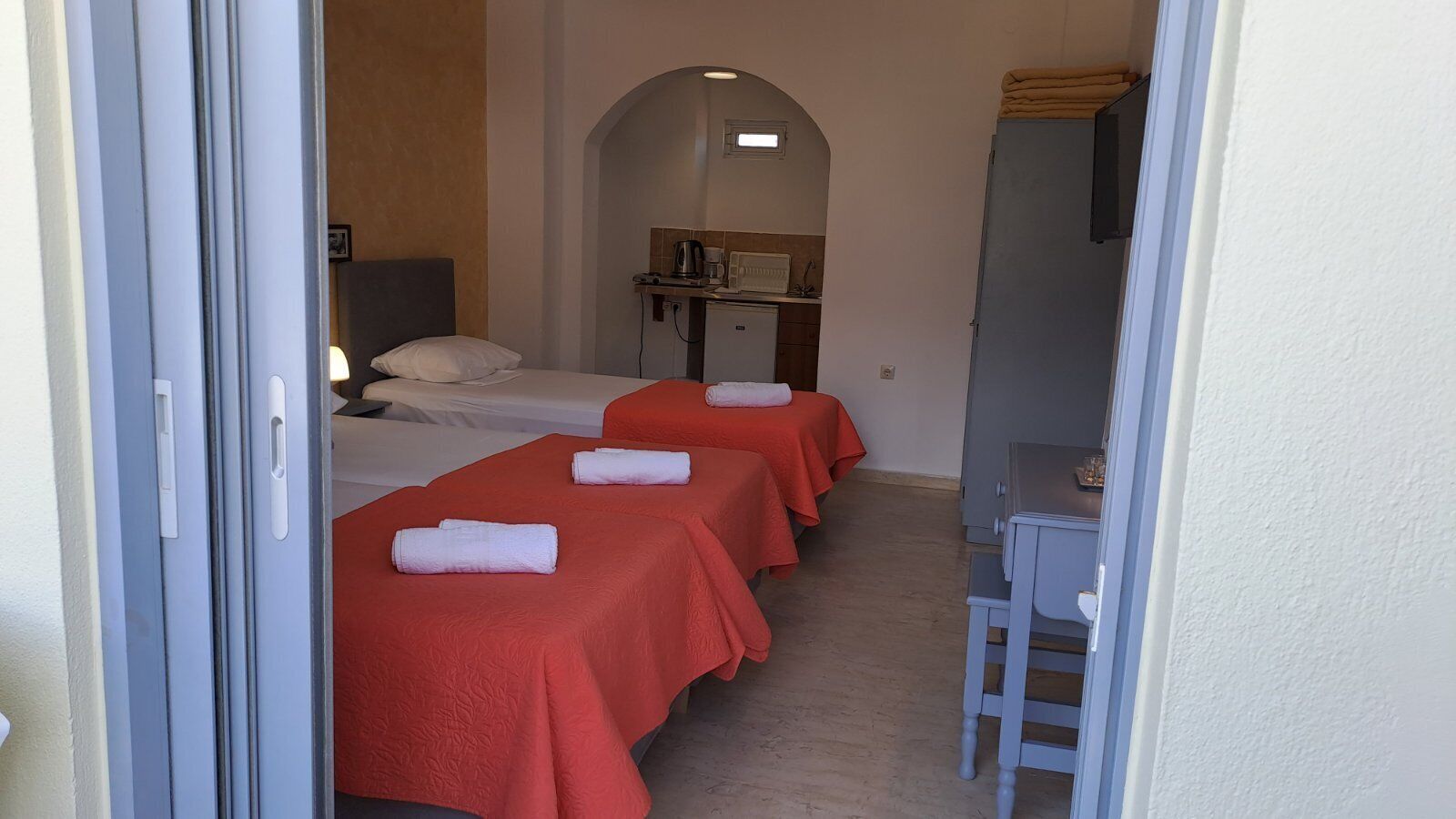 Quarto de hotel com três camas, cada uma com colcha vermelha e toalhas brancas. Dispõe de uma kitchenette com cafeteira e frigorífico.