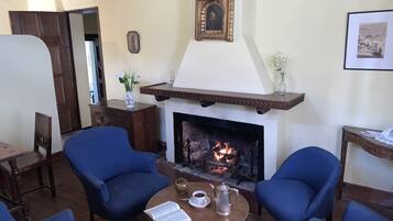 Um quarto aconchegante com lareira, poltronas azuis, uma mesa de madeira com um livro e uma chaleira, e um vaso com flores.