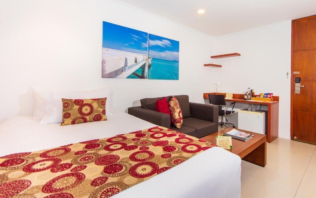 Quarto de hotel com uma cama, um sofá, uma escrivaninha e um pequeno frigorífico. Existem duas pinturas na parede, e uma porta de madeira é visível.
