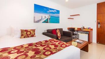 Quarto de hotel com uma cama, um sofá, uma escrivaninha e um pequeno frigorífico. Existem duas pinturas na parede, e uma porta de madeira é visível.