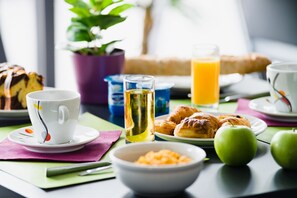 Uma mesa de café da manhã com croissants, maçãs, uma tigela de cereal, um copo de suco de laranja, uma xícara e uma garrafa de azeite.