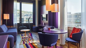 Quarto de hotel moderno com sofá, cadeiras, uma mesa pequena, esteira e vista para a cidade.