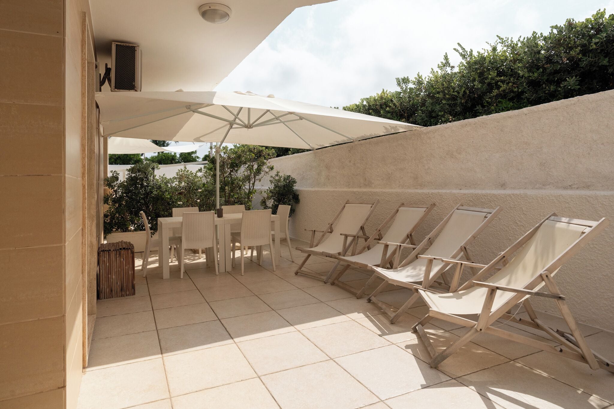 Um pátio externo com mesa e cadeiras, um guarda-sol grande e uma fileira de cadeiras de descanso.