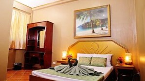 Quarto de hotel com uma cama, uma pintura de uma paisagem praiana, um armário de madeira e duas luminárias de cabeceira.