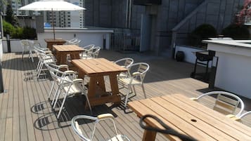 Um terraço ao ar livre com mesas de madeira e cadeiras de metal, um guarda-sol branco e vista para um edifício moderno ao fundo.