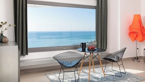 Um quarto com uma janela grande com vista para o mar, uma mesa redonda com duas cadeiras, um vaso com flores secas e um abajur laranja vibrante.