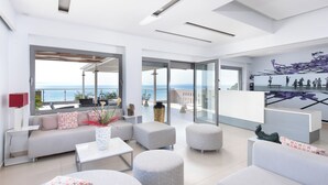 Uma sala de estar moderna com um sofá, pufes e uma mesa de centro. Há uma janela grande com vista para o mar.