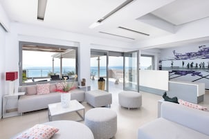 Uma sala de estar moderna com um sofá, pufes e uma mesa de centro. Há uma janela grande com vista para o mar.