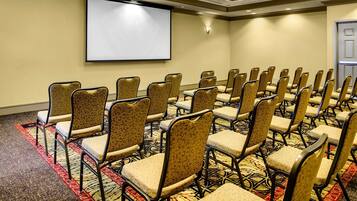 Sala de conferências com fileiras de cadeiras, uma tela de projetor e piso coberto por carpete.