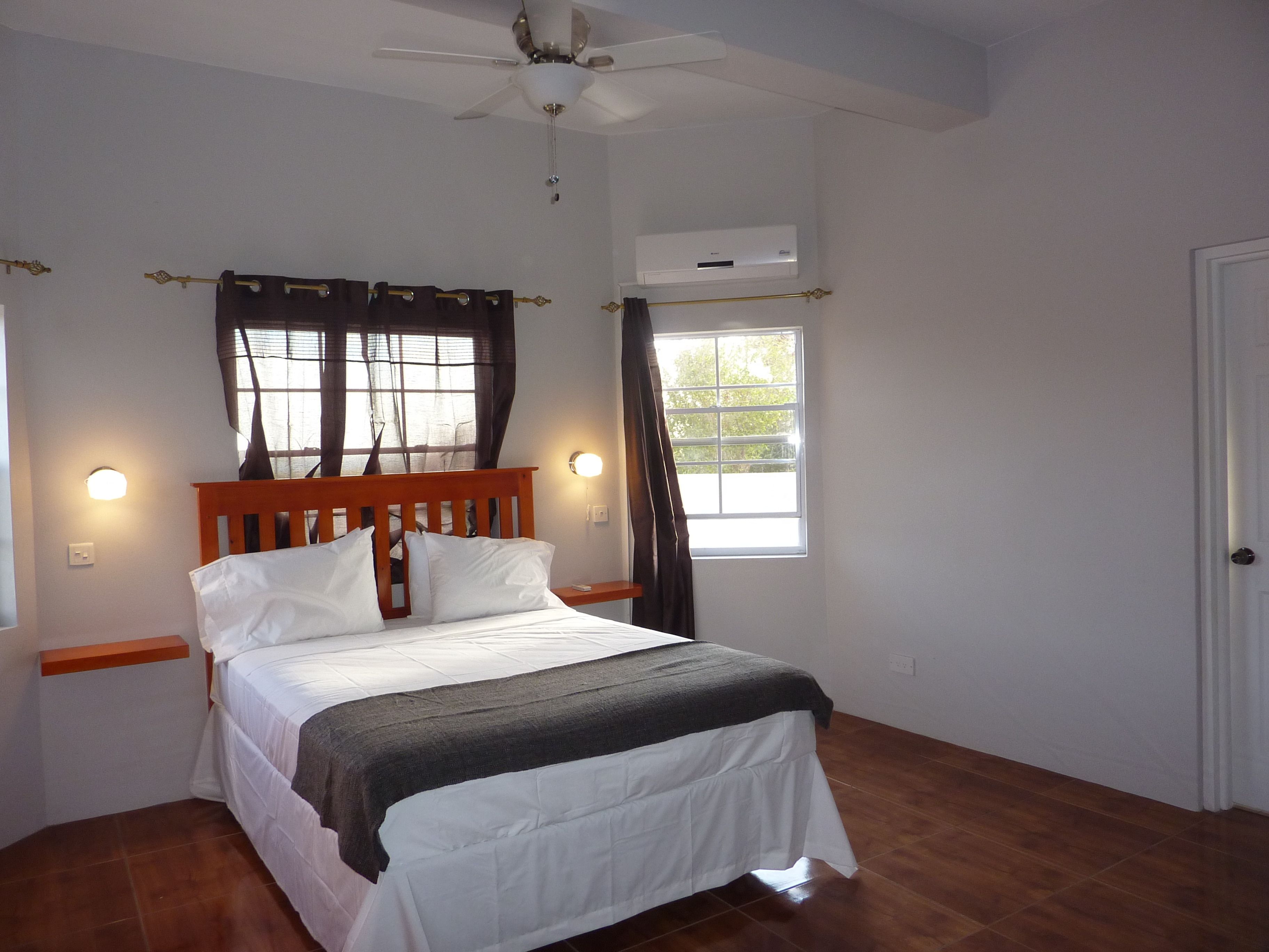 Quarto com cama de madeira, duas mesas de cabeceira, ventoinha no teto, janela com cortinas e ar condicionado.