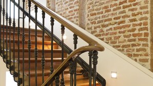 Uma escadaria de madeira com balaústres pretos detalhados e um corrimão, que leva a um quarto com paredes de tijolo aparente.