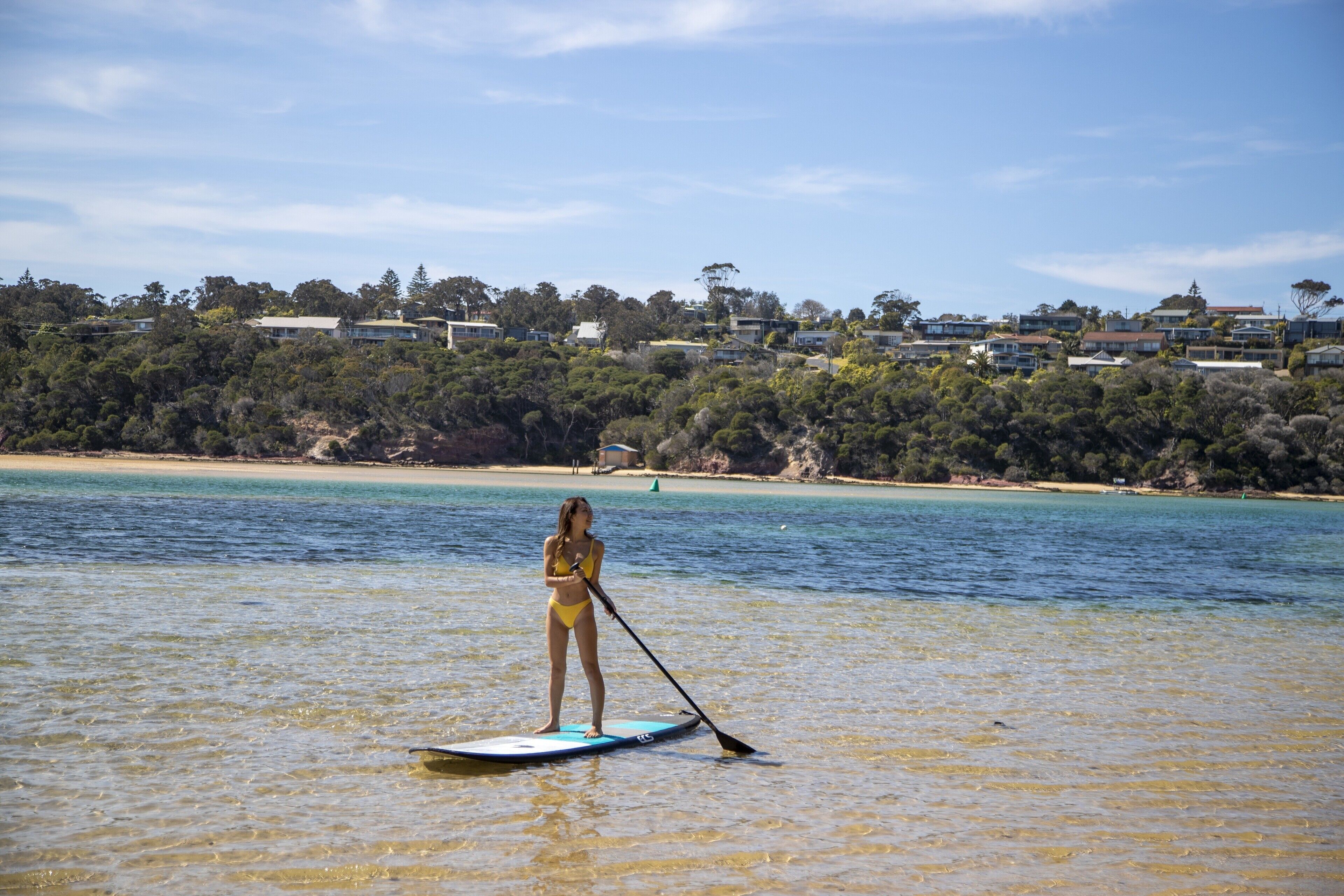 Uma mulher praticando paddleboard num dia claro, com paisagem costeira ao fundo.