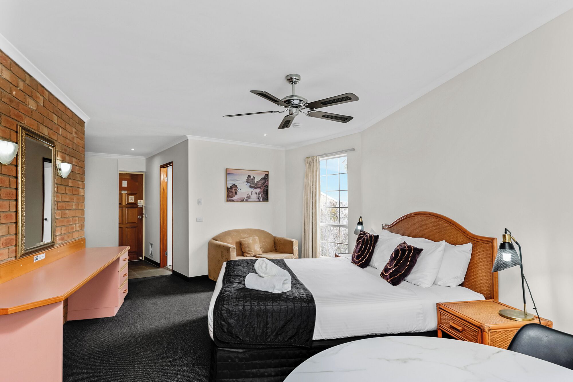 Quarto de hotel com cama, escrivaninha, cadeira, ventoinha no teto e janela.