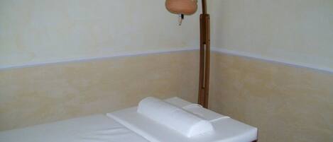 Um quarto pequeno e simples, com uma cama de madeira, um colchão branco e uma mesinha de cabeceira de madeira.