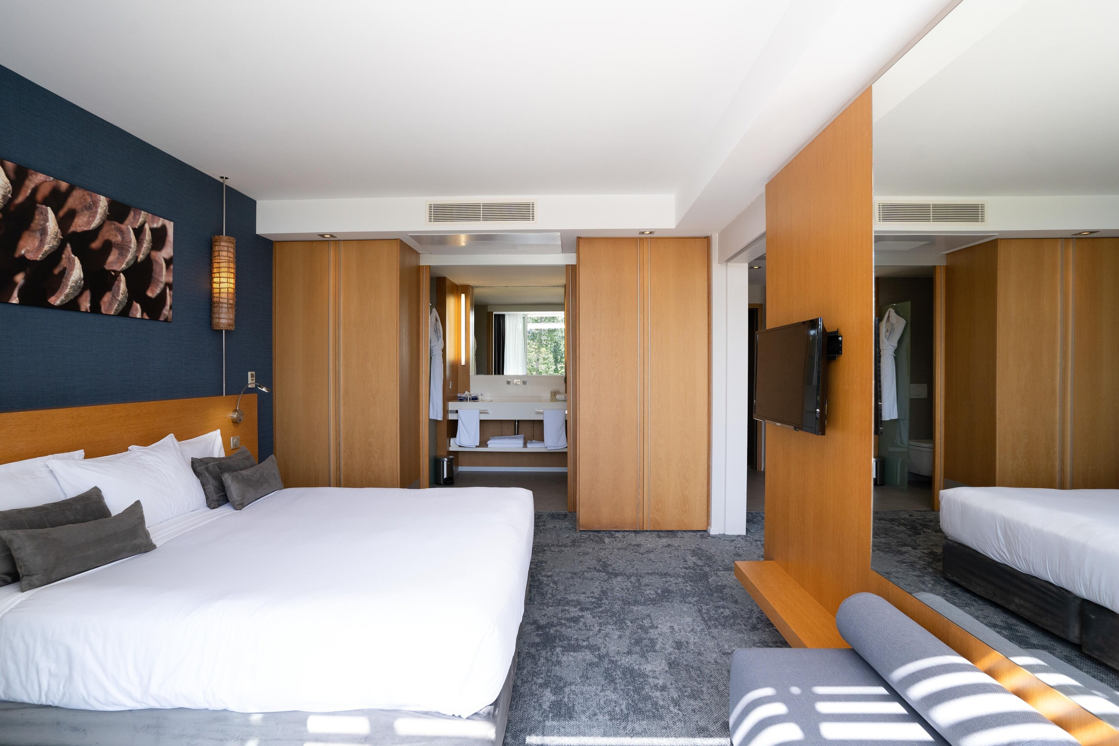 Quarto de hotel moderno com uma cama grande, um sofá, uma mesa pequena e um banheiro visível através de uma porta aberta.