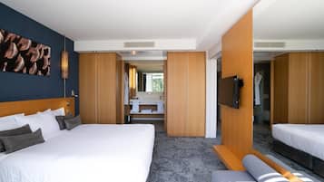 Quarto de hotel moderno com uma cama grande, um sofá, uma mesa pequena e um banheiro visível através de uma porta aberta.