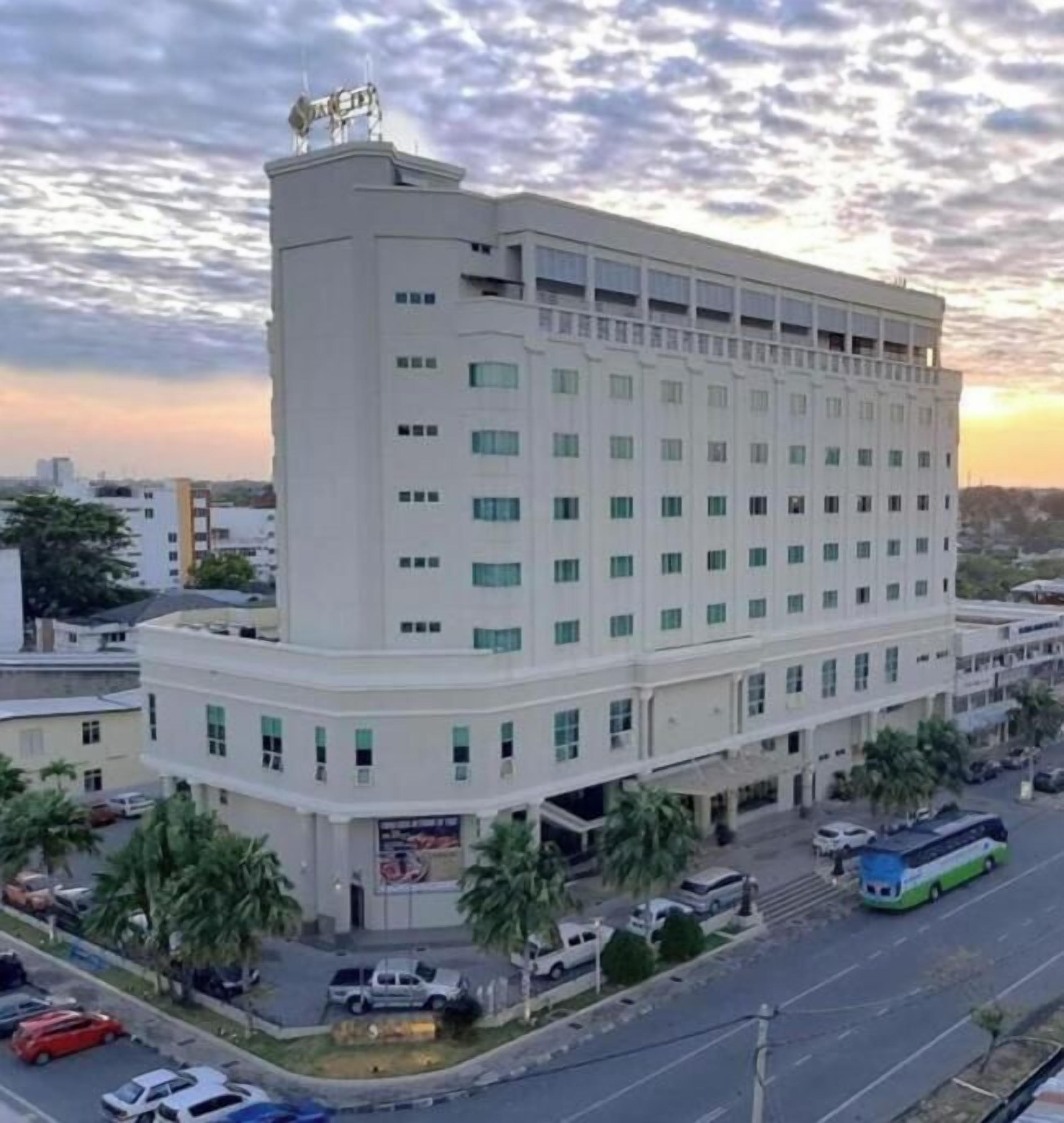 Hotel com vários andares, fachada plana, diversas varandas e área de estacionamento com carros e um ônibus.