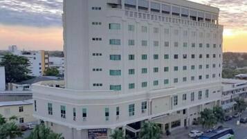 Hotel com vários andares, fachada plana, diversas varandas e área de estacionamento com carros e um ônibus.