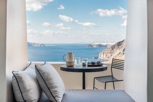 Uma varanda com vista para o mar e falésias, uma mesa com um jarro, copos e um prato de nozes, e um banco acolchoado com almofadas.