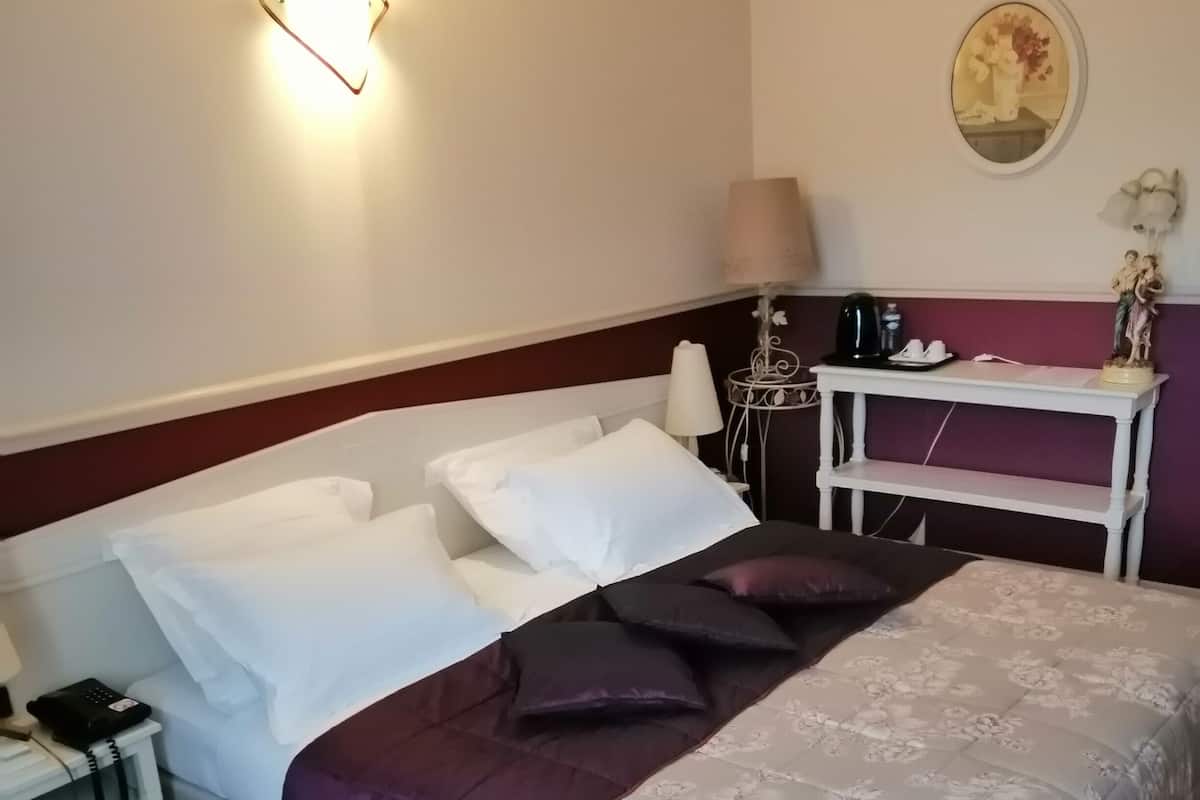 Cama bem arrumada com roupa de cama branca e roxa, mesa de cabeceira com abajur e um pequeno objeto decorativo na parede.