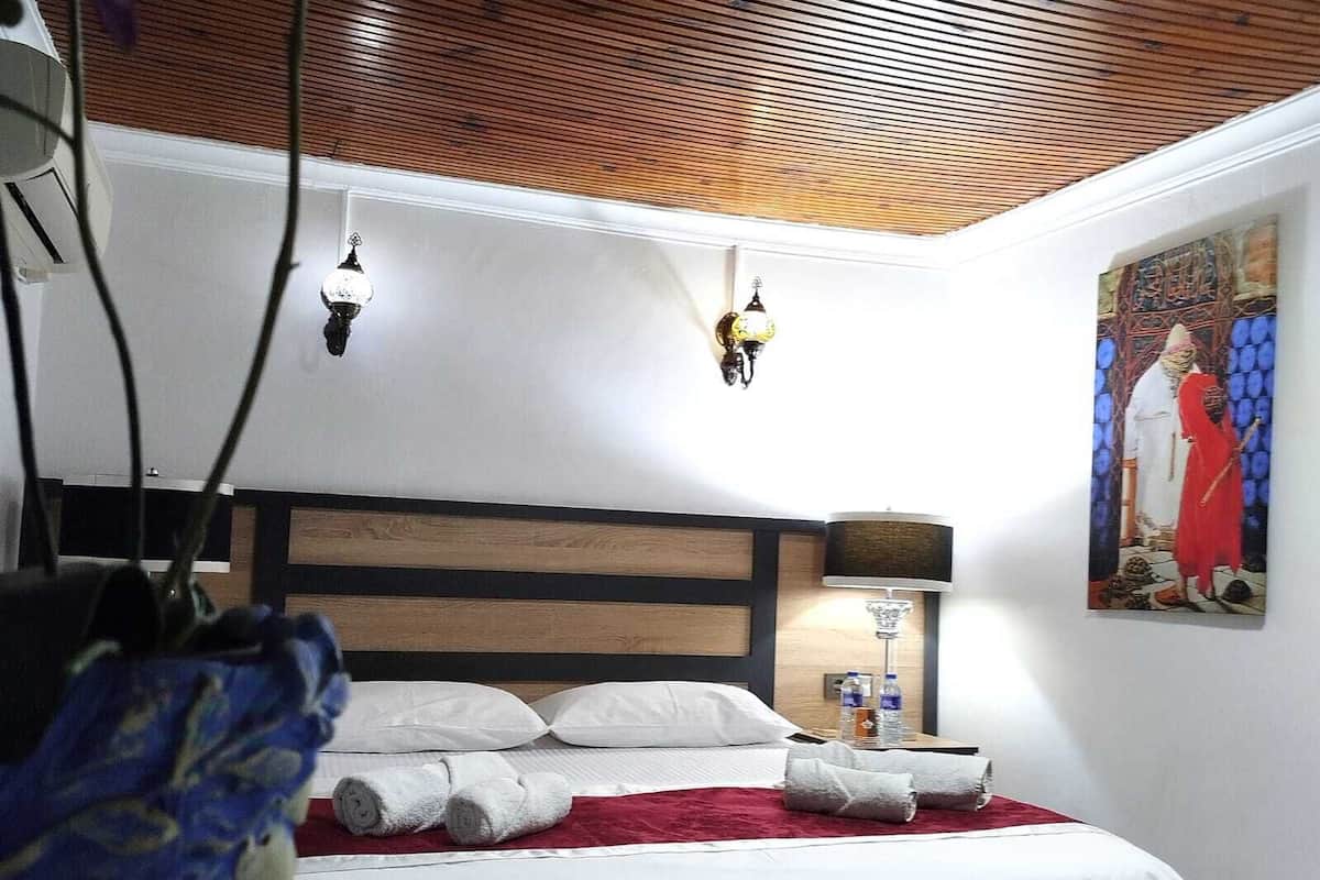 Quarto de hotel com teto de madeira, cama com roupa de cama branca, mesa de cabeceira com abajur e um quadro na parede.