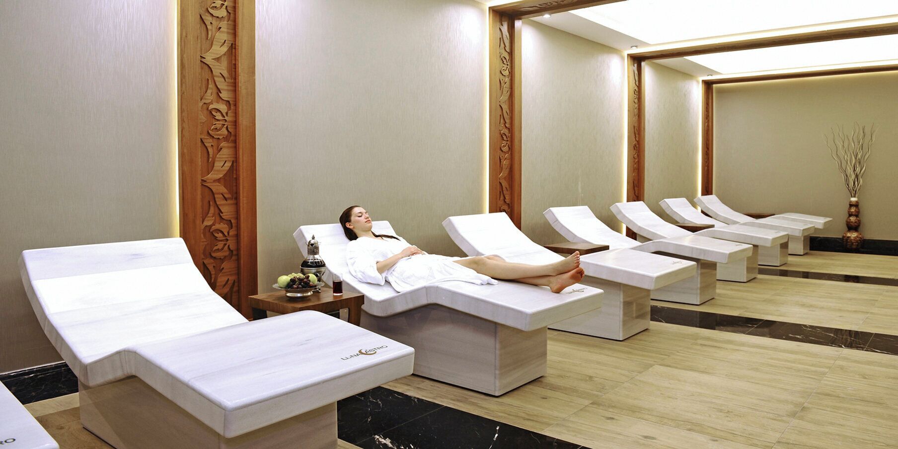 Um quarto de spa com várias mesas de massagem dispostas em linha, uma pessoa deitada em uma delas, e uma mesinha com uma cesta de frutas e uma garrafa.