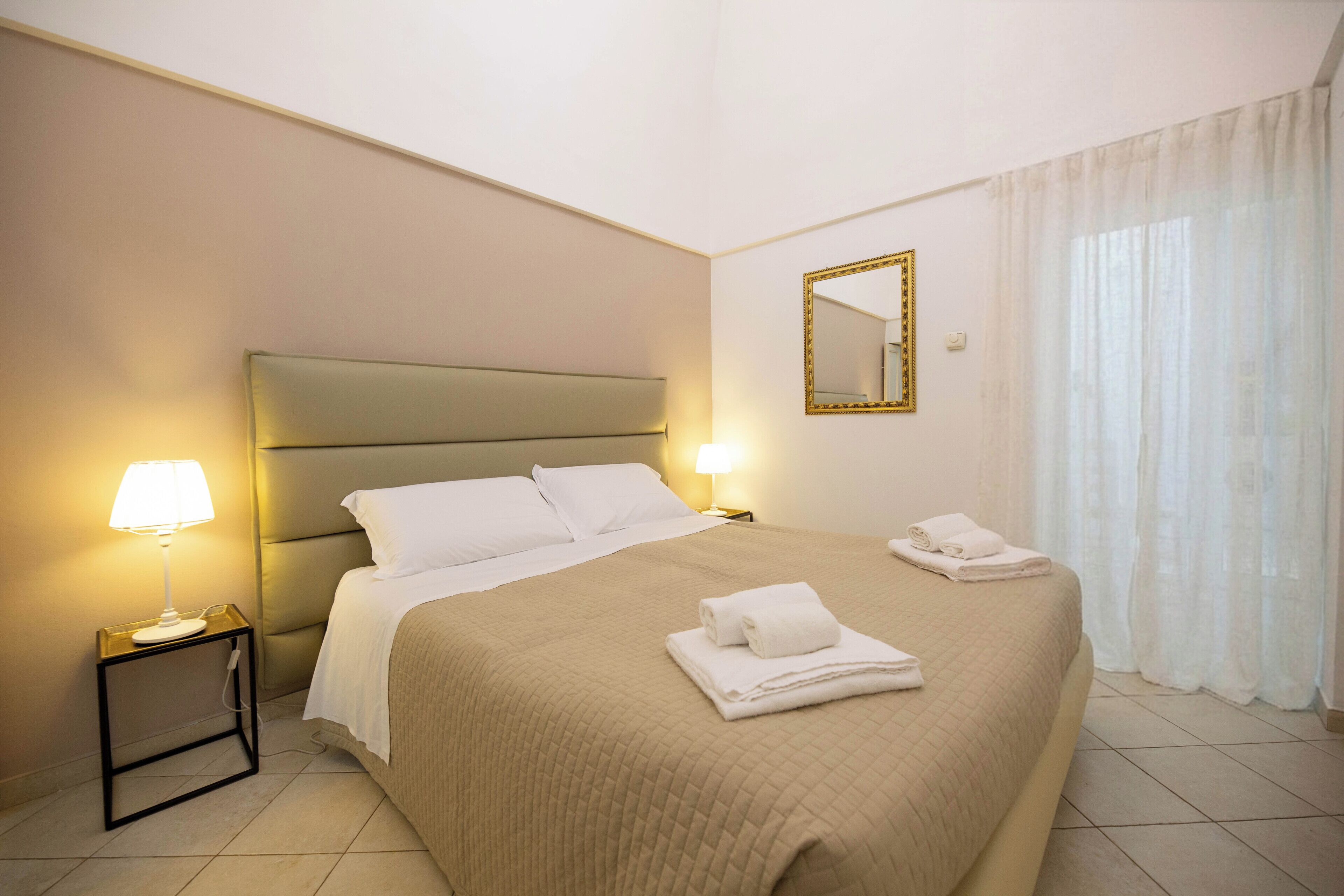 Quarto de hotel com cama grande, abajur, espelho e janela com cortinas.