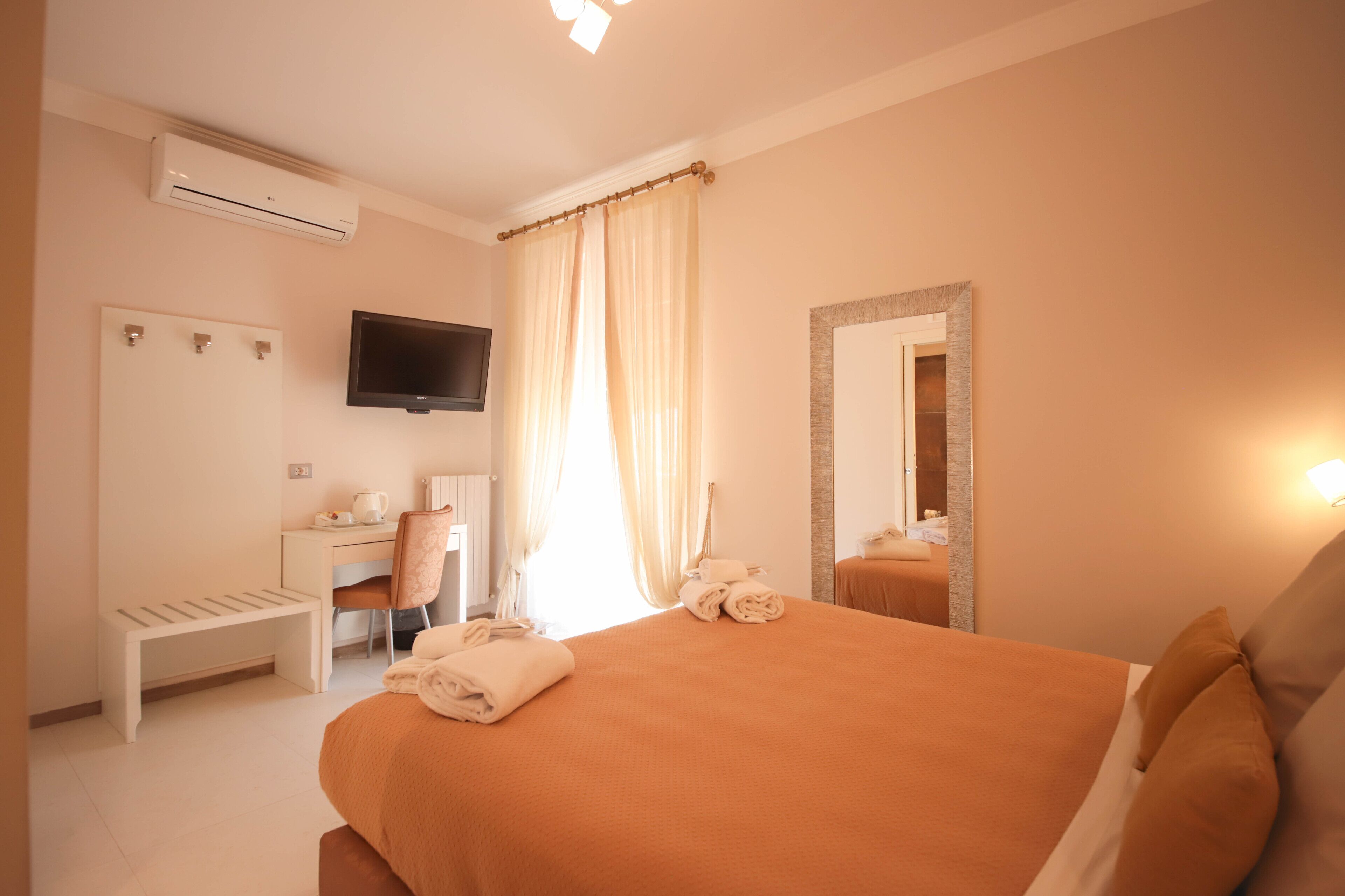 Quarto de hotel com cama, escrivaninha, cadeira, televisão e espelho.
