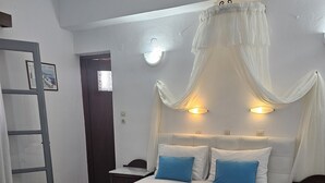 Uma cama bem arrumada com colcha azul estampada e duas almofadas, uma mesa de cabeceira com abajur, e uma porta com cortina.