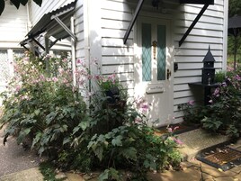 Uma casa branca com porta verde, comedouro de pássaros e flores cor-de-rosa no jardim.