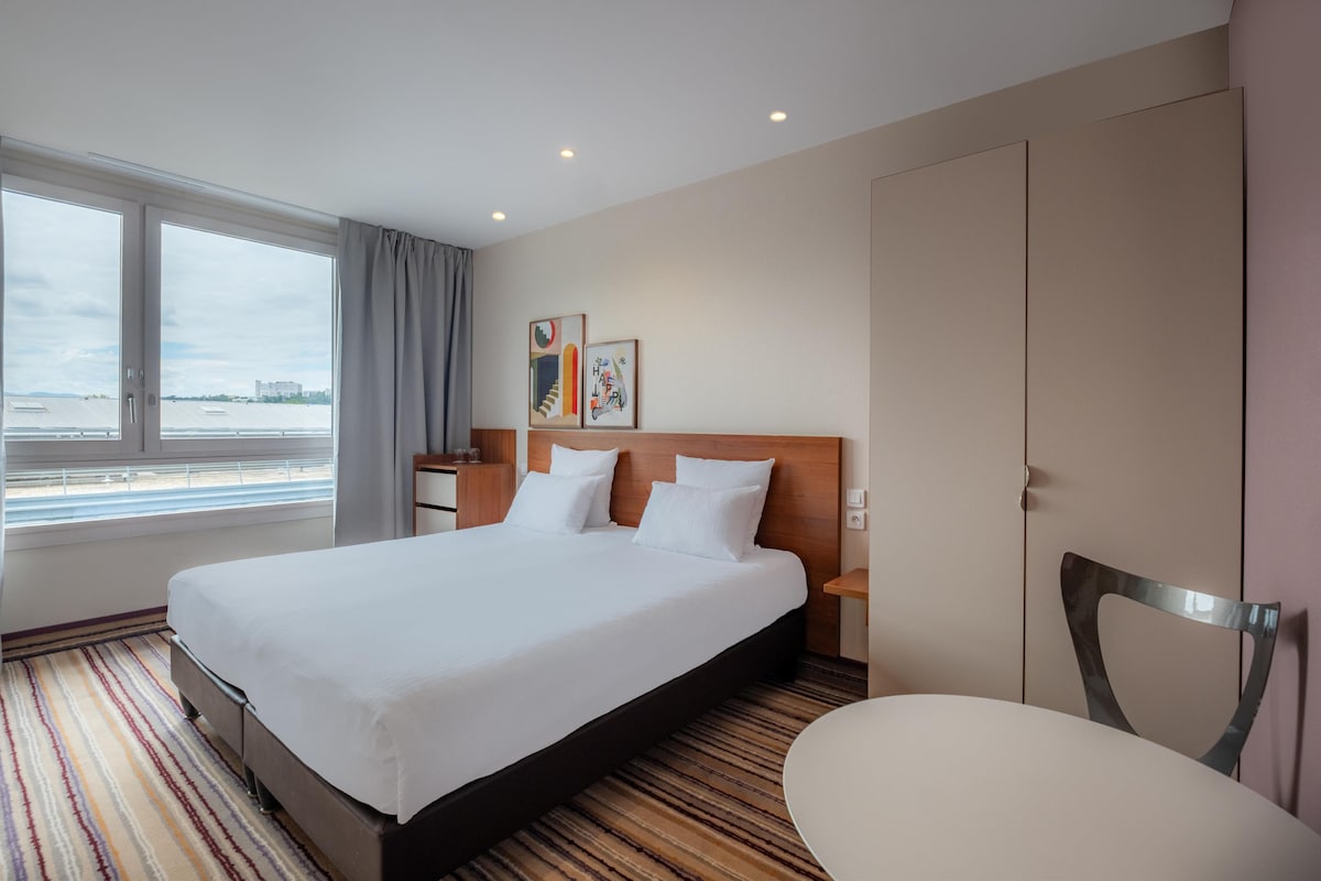 Quarto de hotel com uma cama grande, uma cadeira, uma mesa e uma janela com vista para a água.
