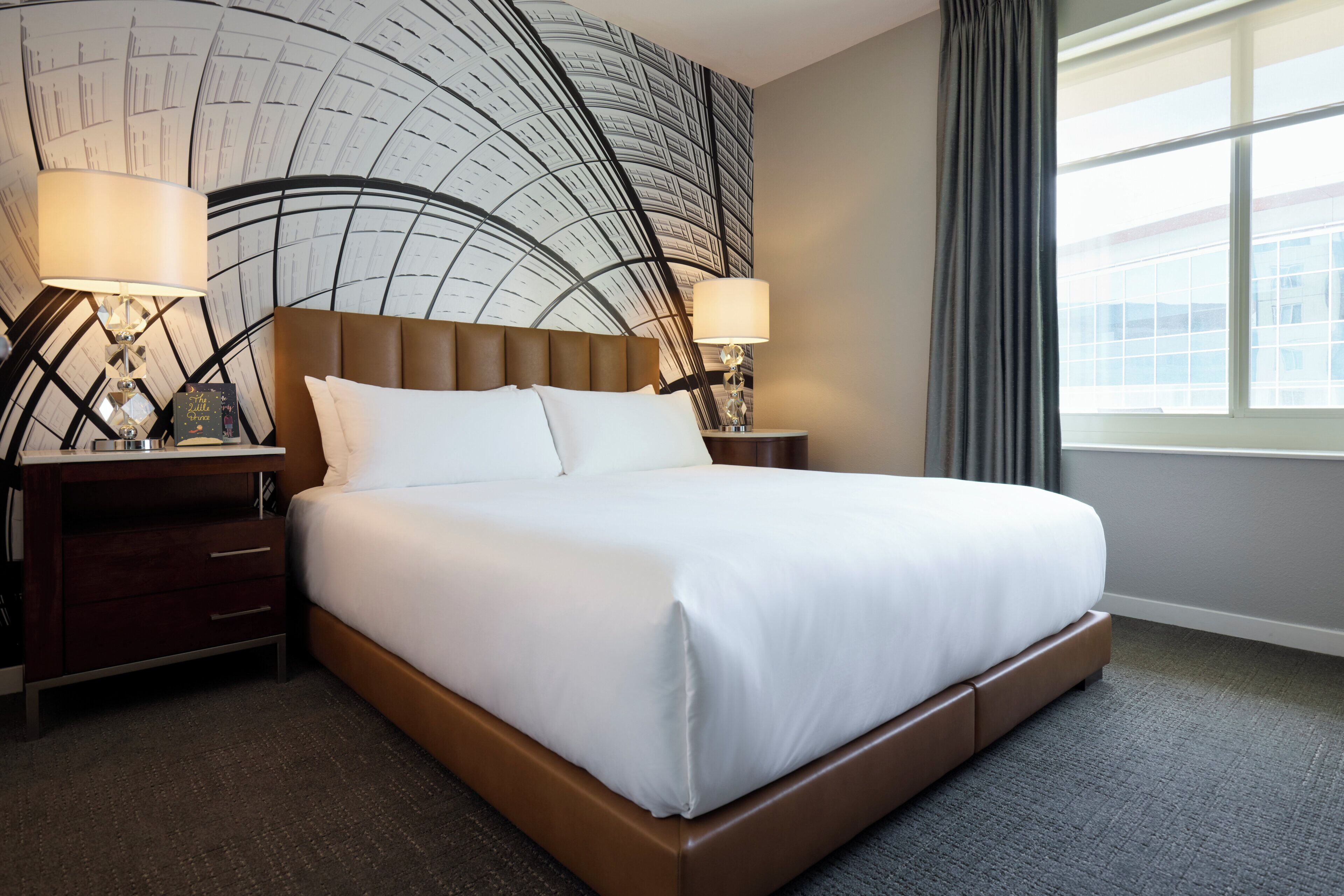 Quarto de hotel com uma cama grande, duas mesas de cabeceira, uma parede com padrão geométrico e uma janela com cortinas.