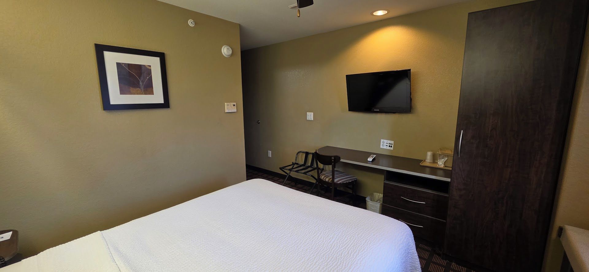 Quarto de hotel com cama, escrivaninha, cadeira, televisão e guarda-roupa.