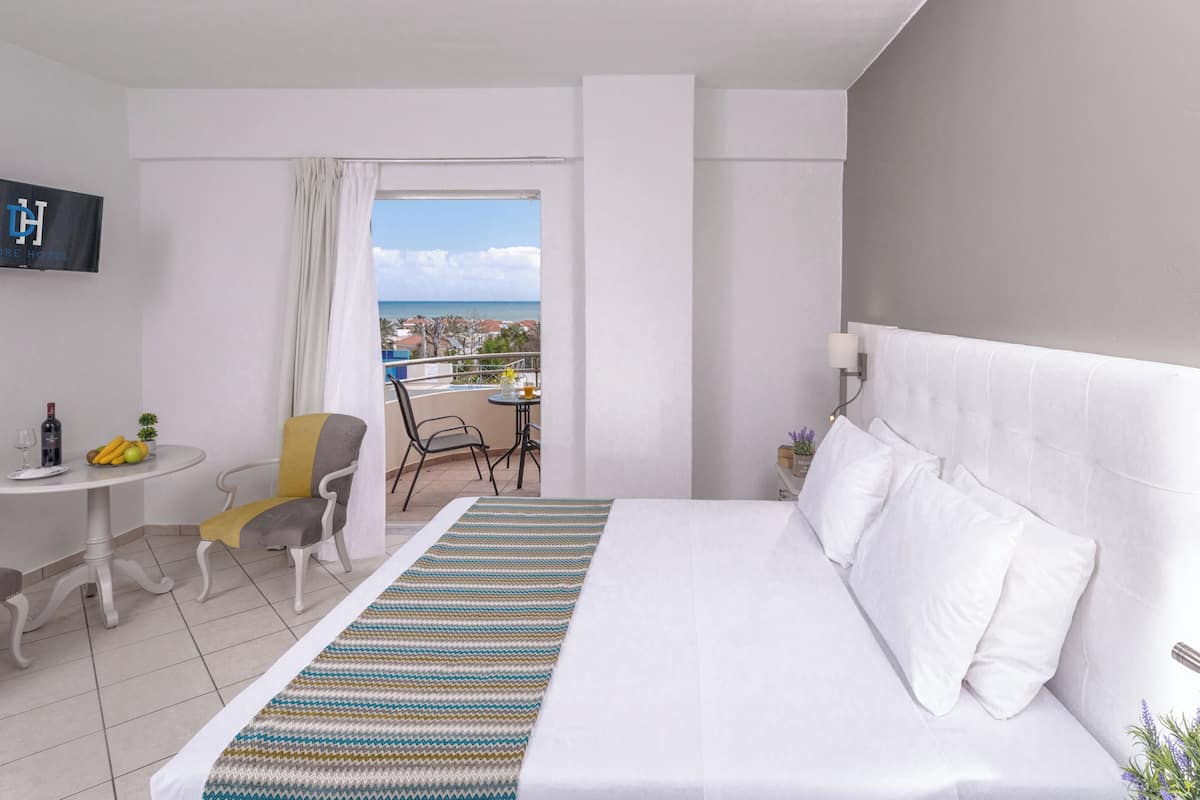 Quarto de hotel com cama, televisão, varanda com vista e uma pequena mesa com frutas e uma garrafa.