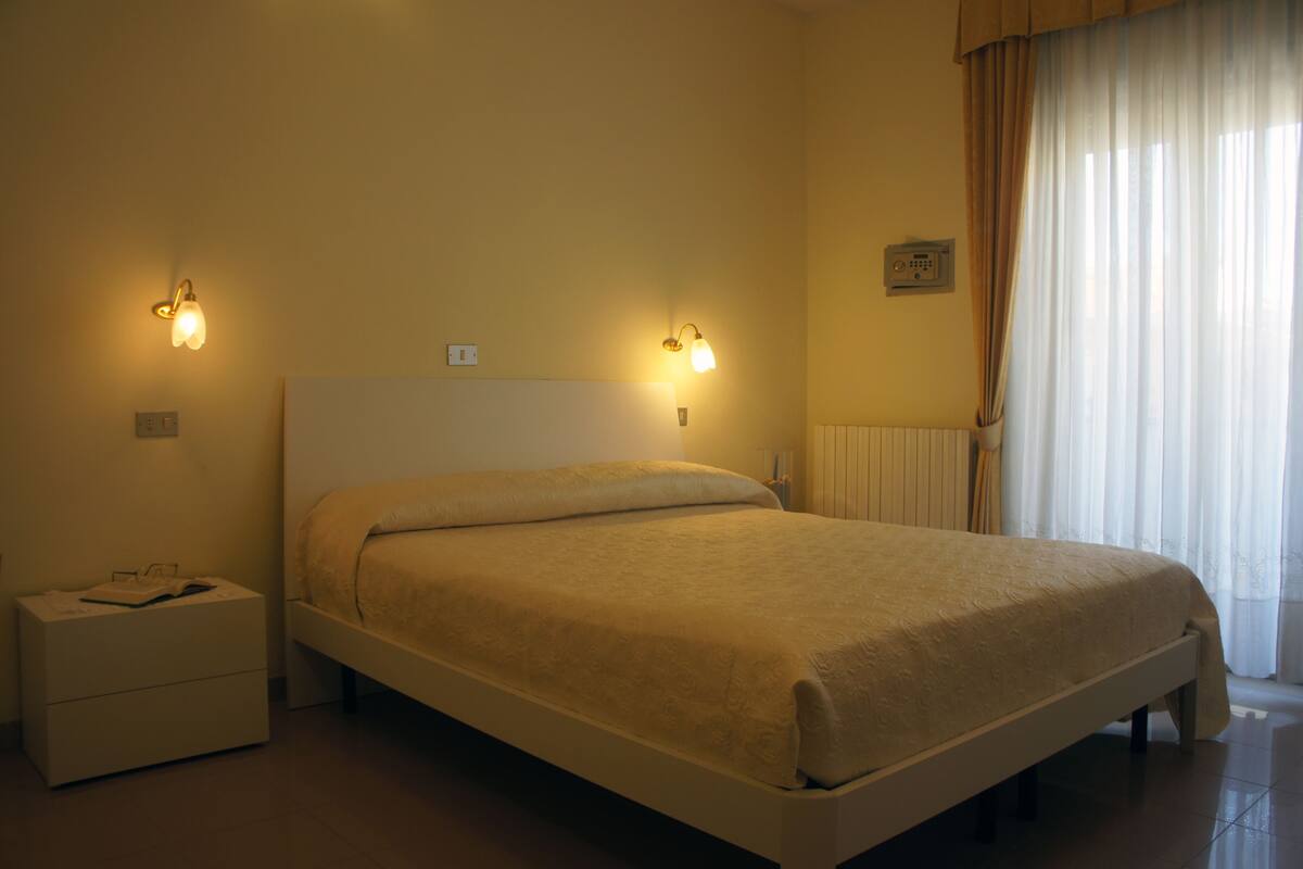 Quarto de hotel com uma cama de solteiro, mesas de cabeceira e luminárias fixadas na parede.