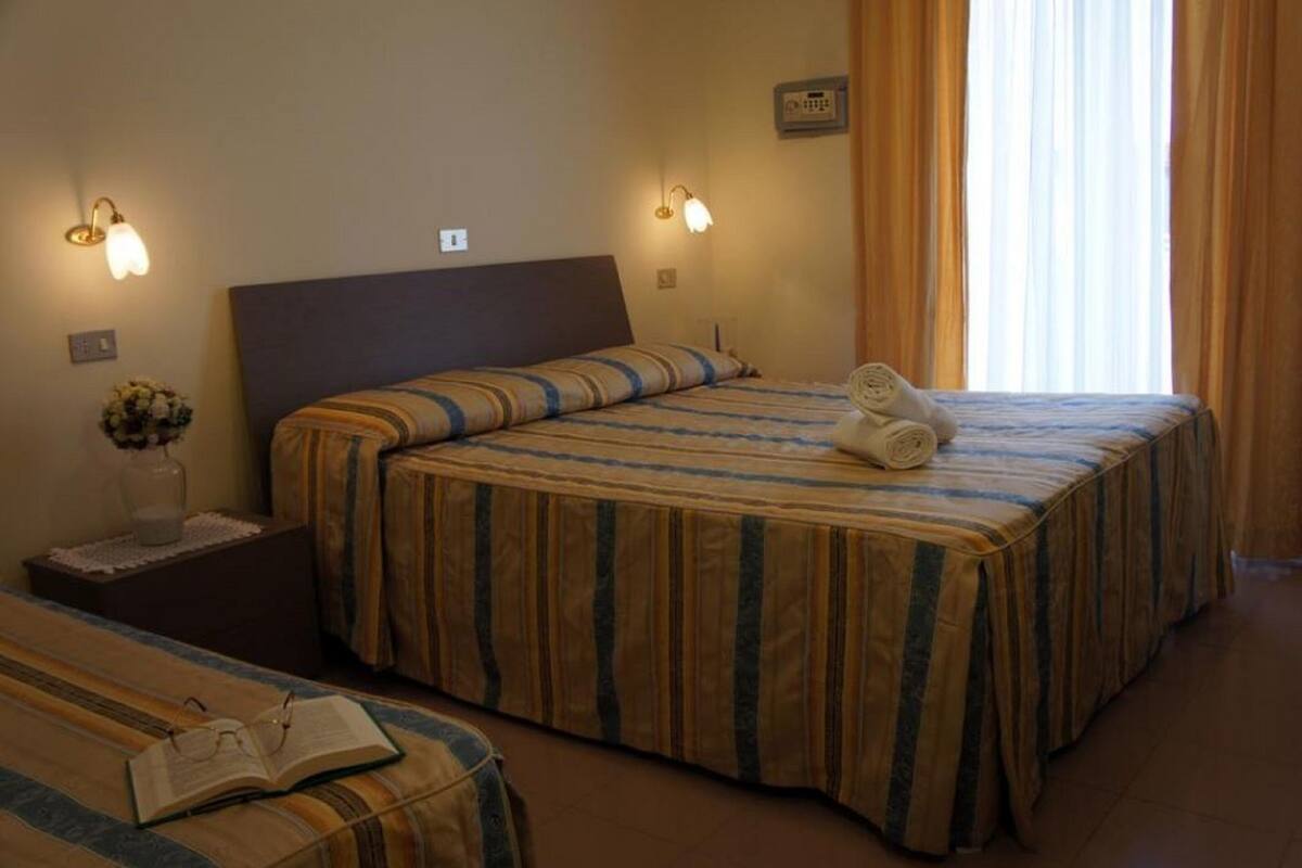Quarto de hotel com uma cama de solteiro, mesa de cabeceira com abajur, pequena mesa com um vaso de flores e uma janela com cortinas.