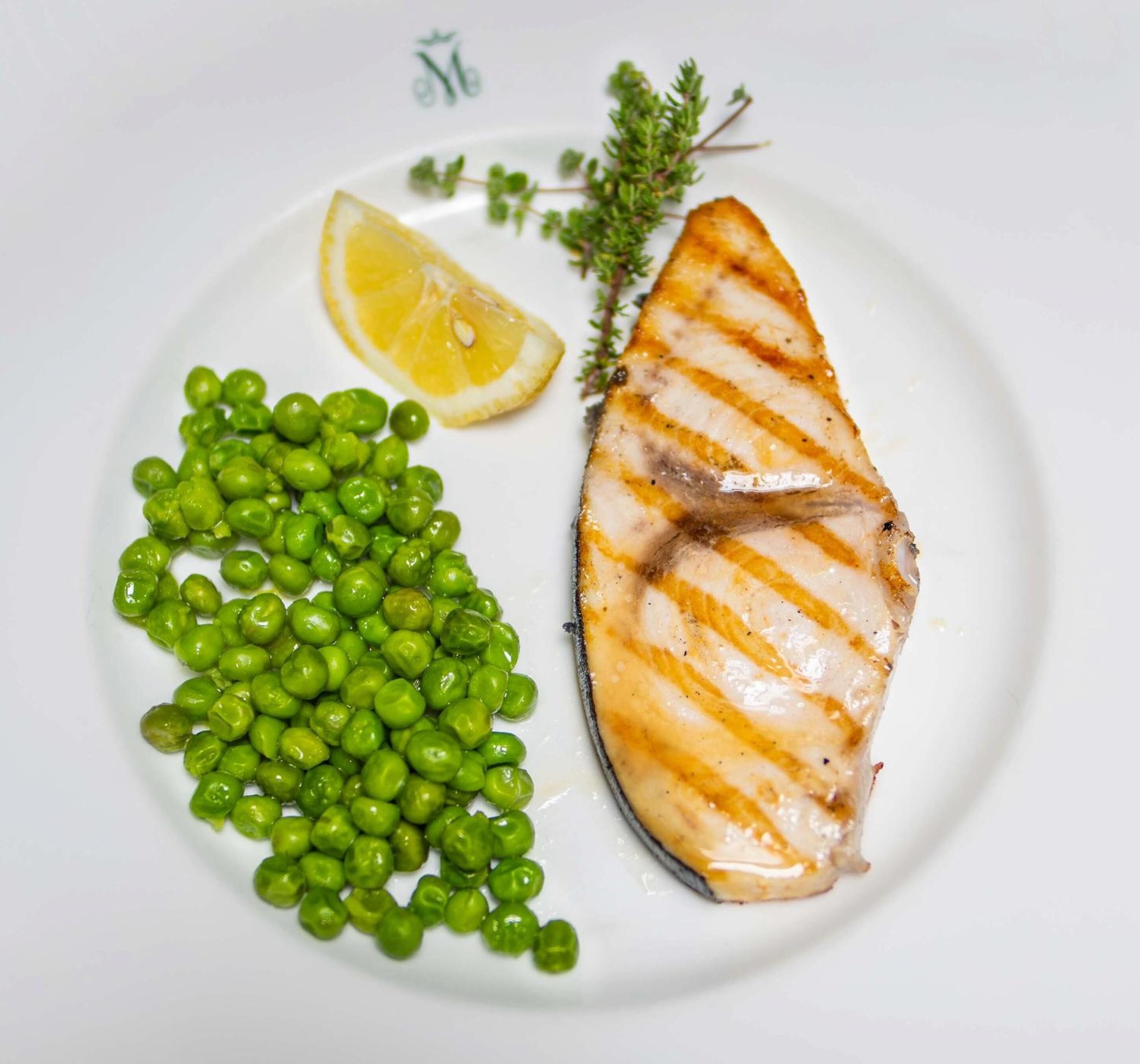 Um prato com peixe grelhado, ervilhas verdes, uma fatia de limão e um raminho de tomilho.