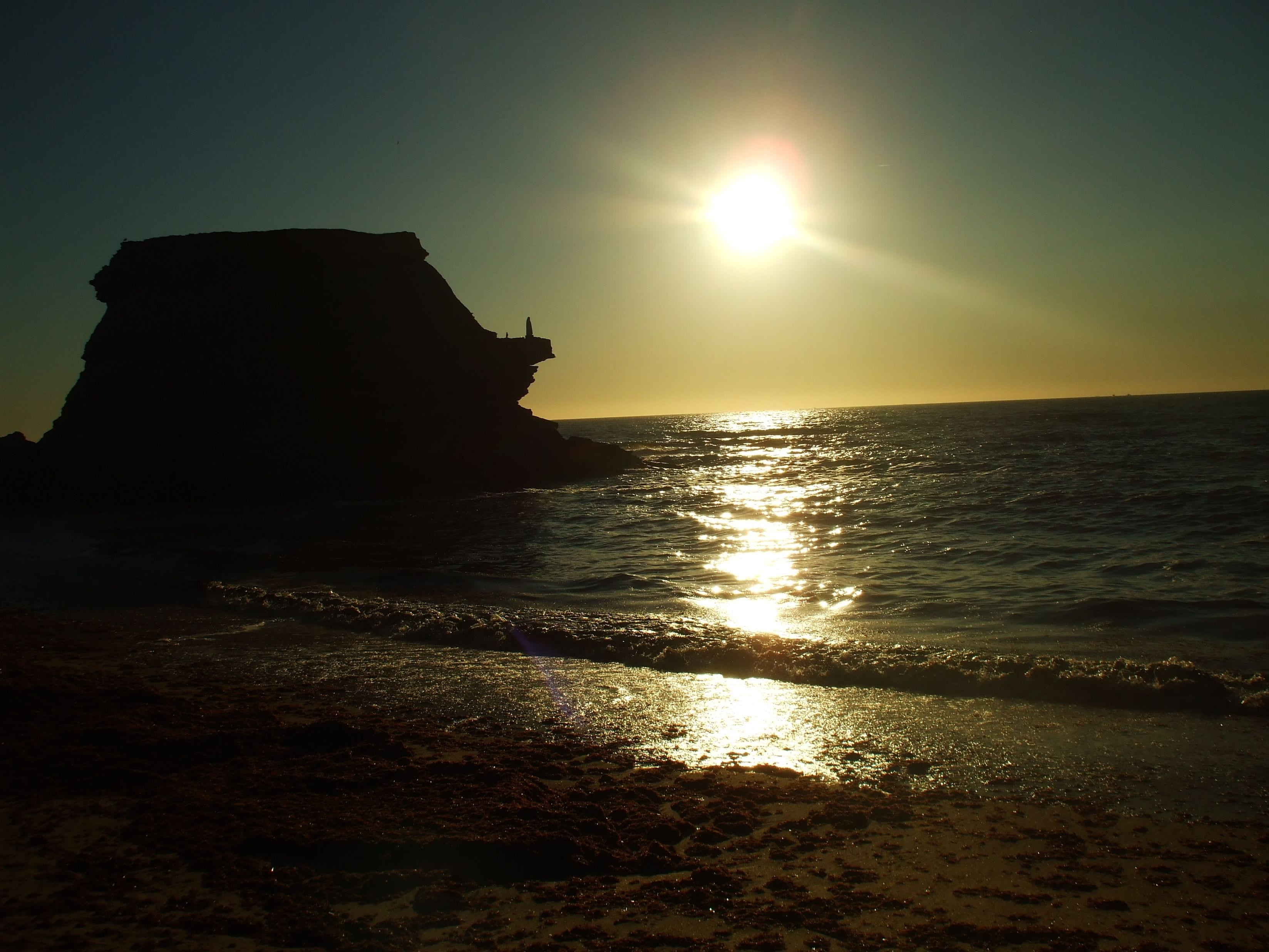 Uma praia com um afloramento rochoso e o sol refletindo na água.