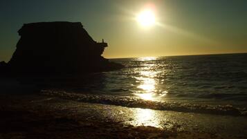 Uma praia com um afloramento rochoso e o sol refletindo na água.