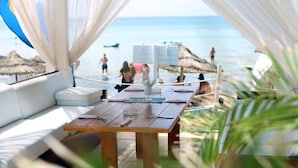 Uma área de refeições à beira-mar com mesa de madeira, almofadas brancas e vista para o mar e pessoas na praia.
