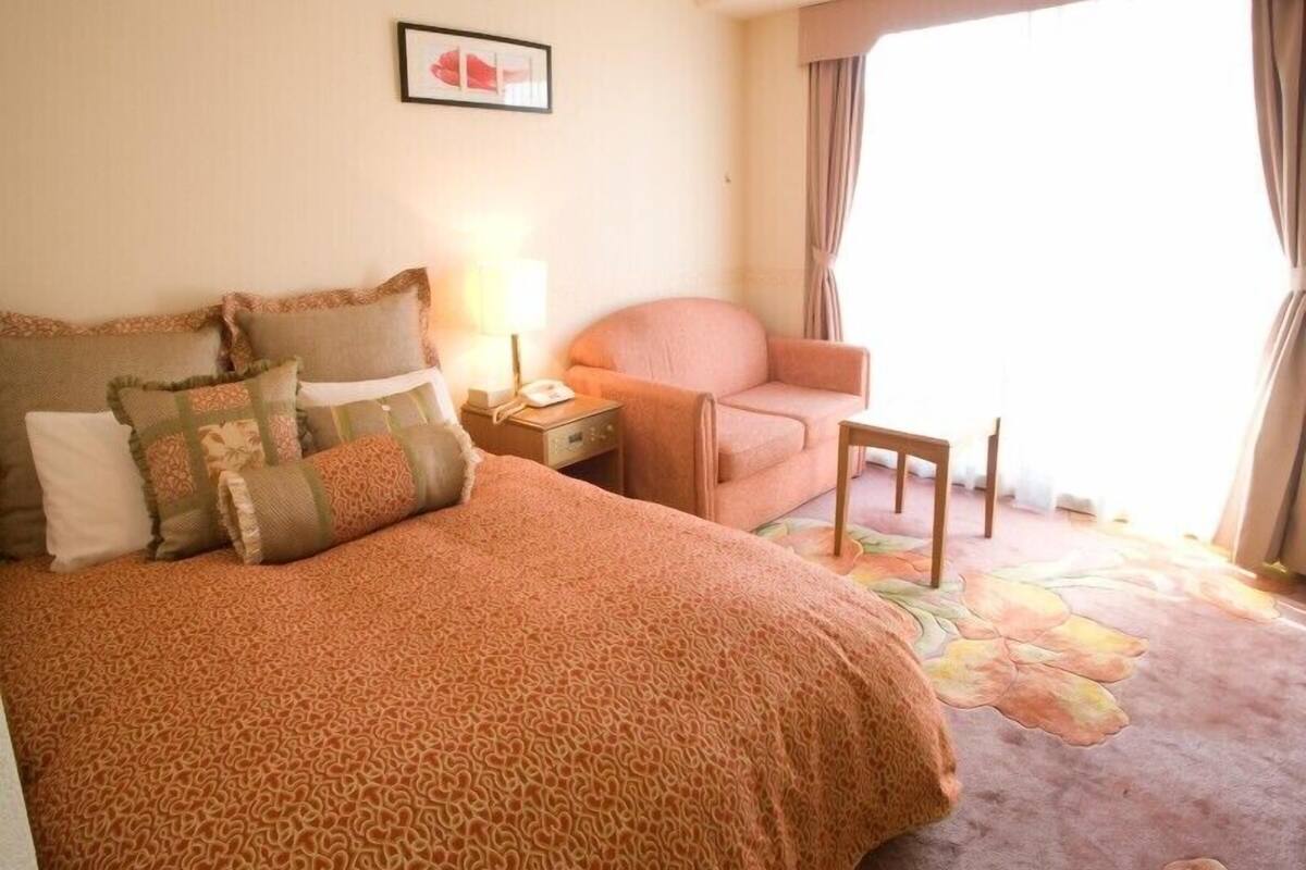 Quarto de hotel com cama, cadeira, mesa de cabeceira e janela com cortinas.
