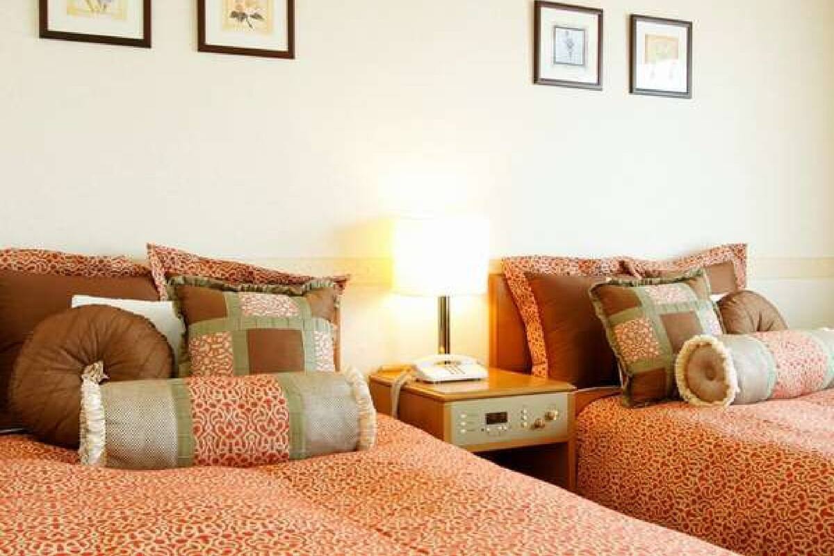 Quarto de hotel com duas camas, cada uma com roupa de cama estampada e várias almofadas. Há quadros nas paredes e uma mesa de cabeceira com abajur.