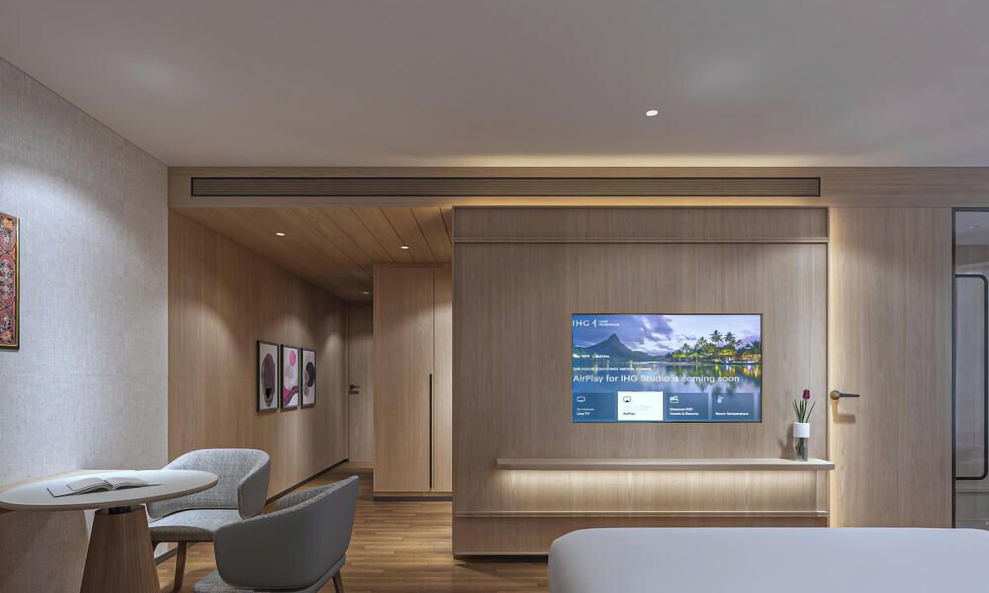 Quarto de hotel moderno com uma grande TV de tela plana na parede, uma cama, um espaço de estar com cadeiras e piso de madeira.