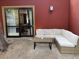 Uma área de estar ao ar livre com assentos brancos, mesa de vime e sofá, porta de vidro deslizante e luminária fixa na parede.