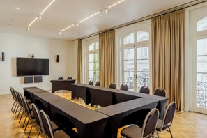 Uma sala de conferências com uma mesa comprida, cadeiras, uma TV de tela plana e janelas amplas com cortinas.