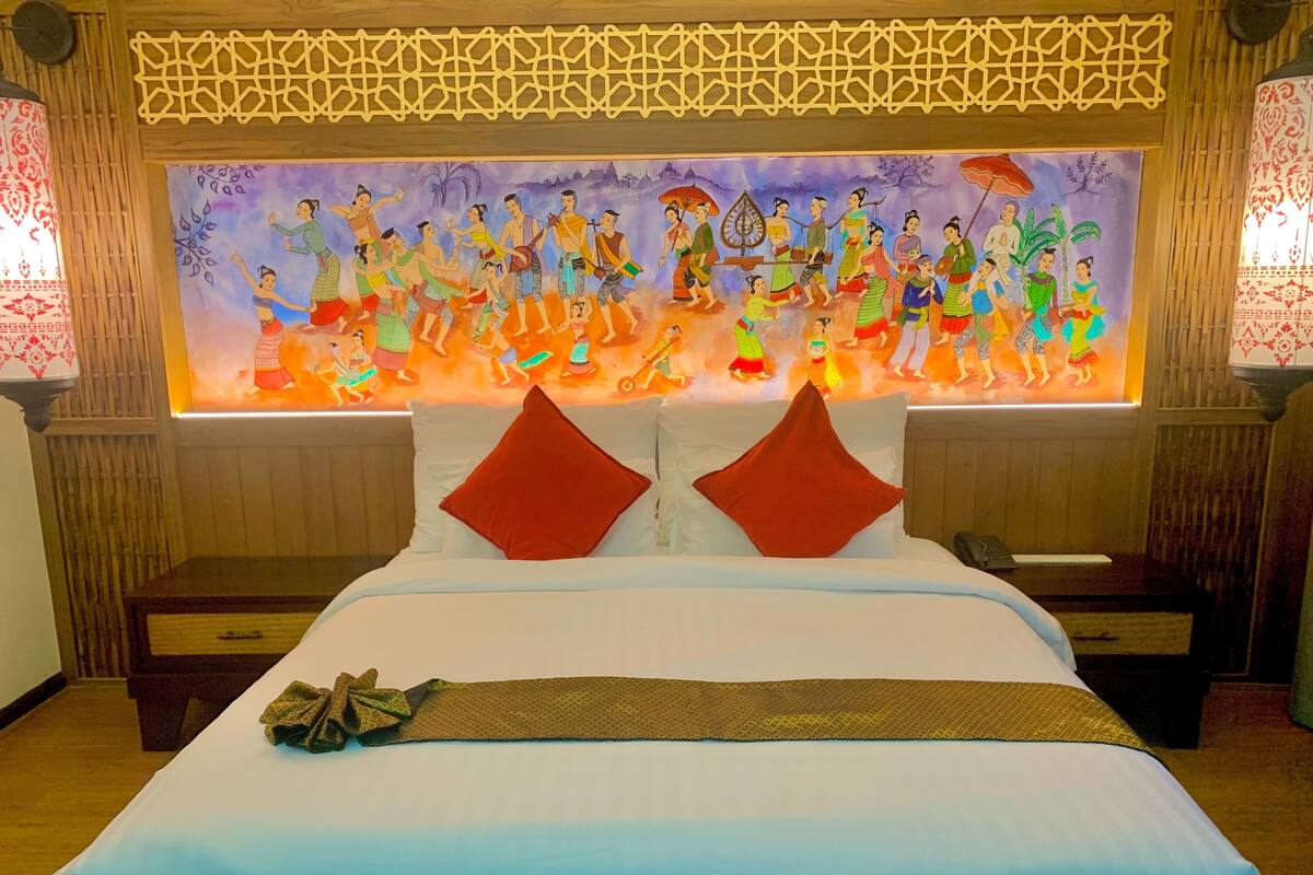 Quarto de hotel com um grande mural na parede, uma cama com roupa de cama branca e almofadas vermelhas, e duas mesas de cabeceira com luminárias.