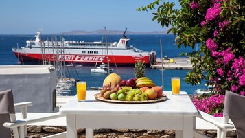 Uma mesa com frutas e um copo de suco de laranja, com vista para uma marina com barcos e um ferry.